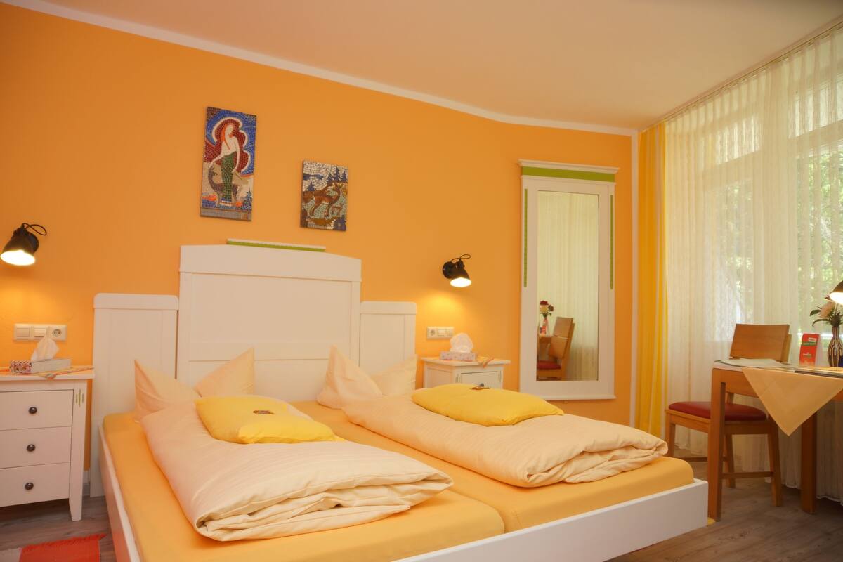 Uma cama de casal com duas almofadas, uma mesinha de cabeceira com abajur, uma pequena mesa com cadeira e uma janela com cortinas.