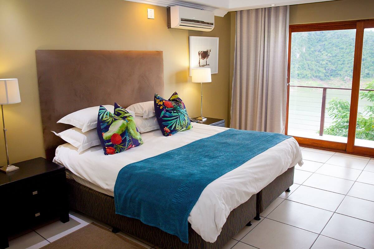 Quarto de hotel com cama, mesas de cabeceira, abajur, ar condicionado, janela com cortinas e vista para o verde.