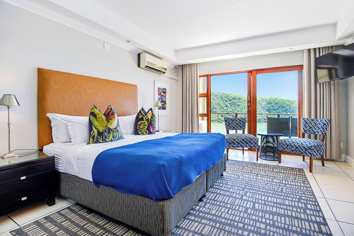 Quarto de hotel com uma cama grande, uma varanda com cadeiras e uma televisão.