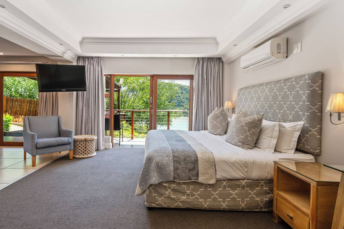 Quarto de hotel moderno com uma cama grande, televisão, varanda com vista para vegetação e uma mesa de cabeceira de madeira.