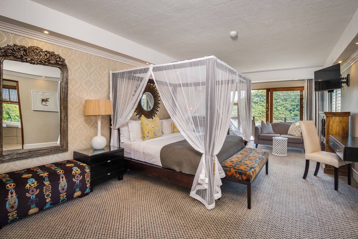 Um quarto com cama de dossel, área de estar com sofá e poltronas, televisão e uma janela grande com vista para o exterior.