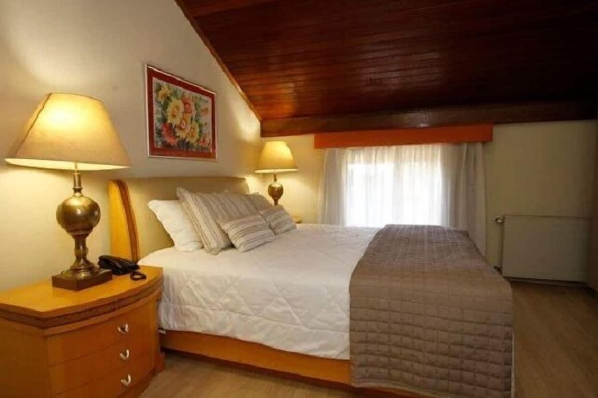 Quarto com cama, duas luminárias de cabeceira, mesa de apoio, um quadro na parede e uma janela com cortinas.