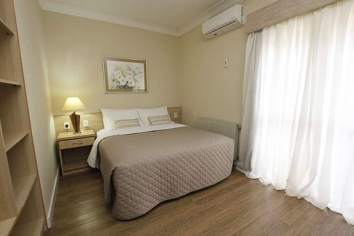 Quarto de hotel com uma cama, mesa de cabeceira, luminária, um quadro na parede e janela com cortinas.
