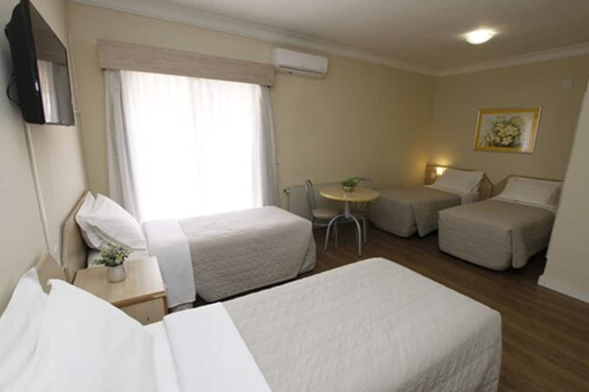 Quarto de hotel com duas camas de solteiro, uma mesa pequena, uma cadeira e uma janela com cortinas.