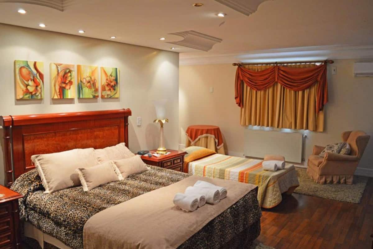 Quarto de hotel com uma cama grande, duas camas menores, um espaço de estar com uma poltrona e obras de arte na parede.
