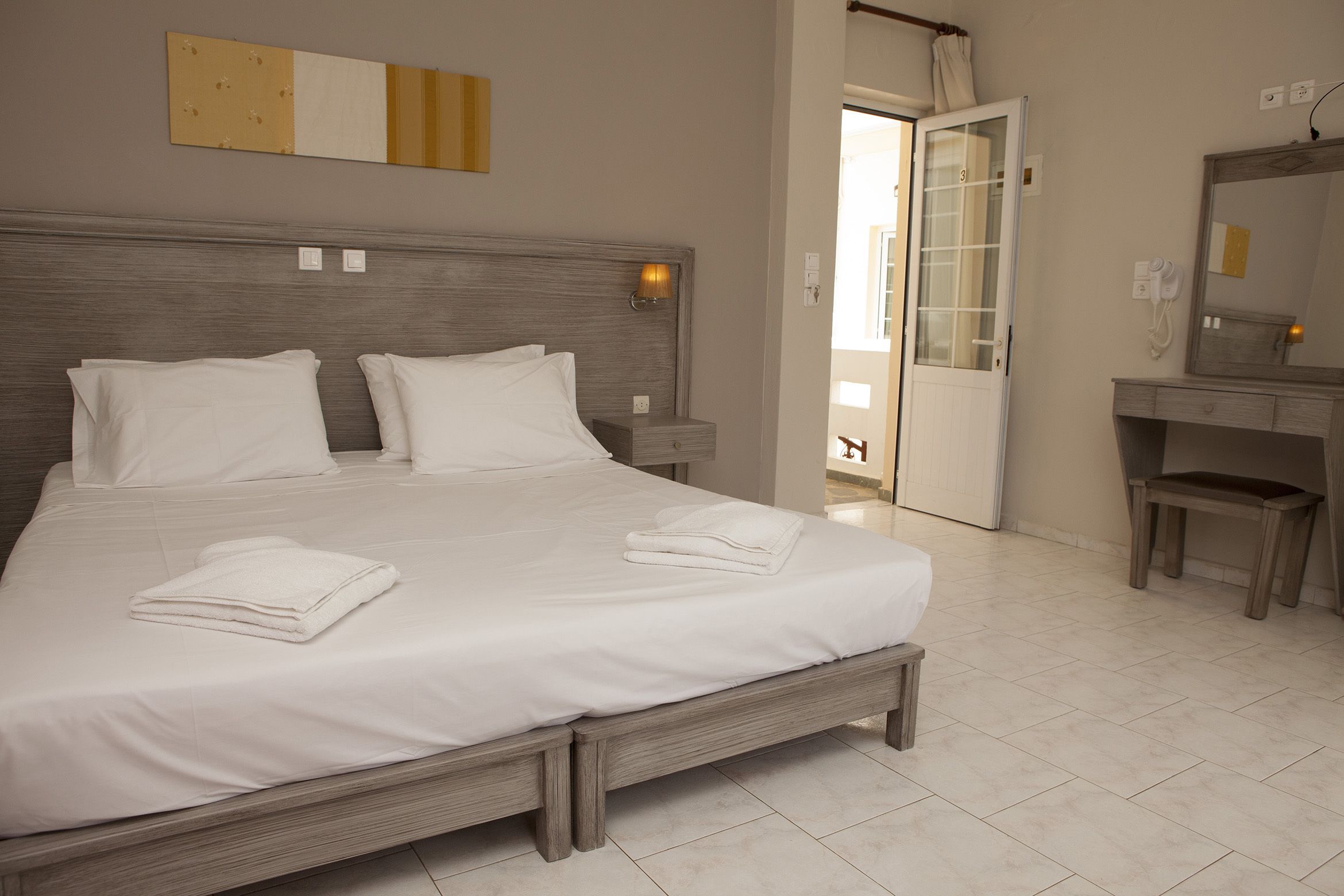 Cama bem arrumada com lençóis brancos e toalhas dobradas, cabeceira de madeira, luminária de cabeceira e banheiro com espelho e toucador.