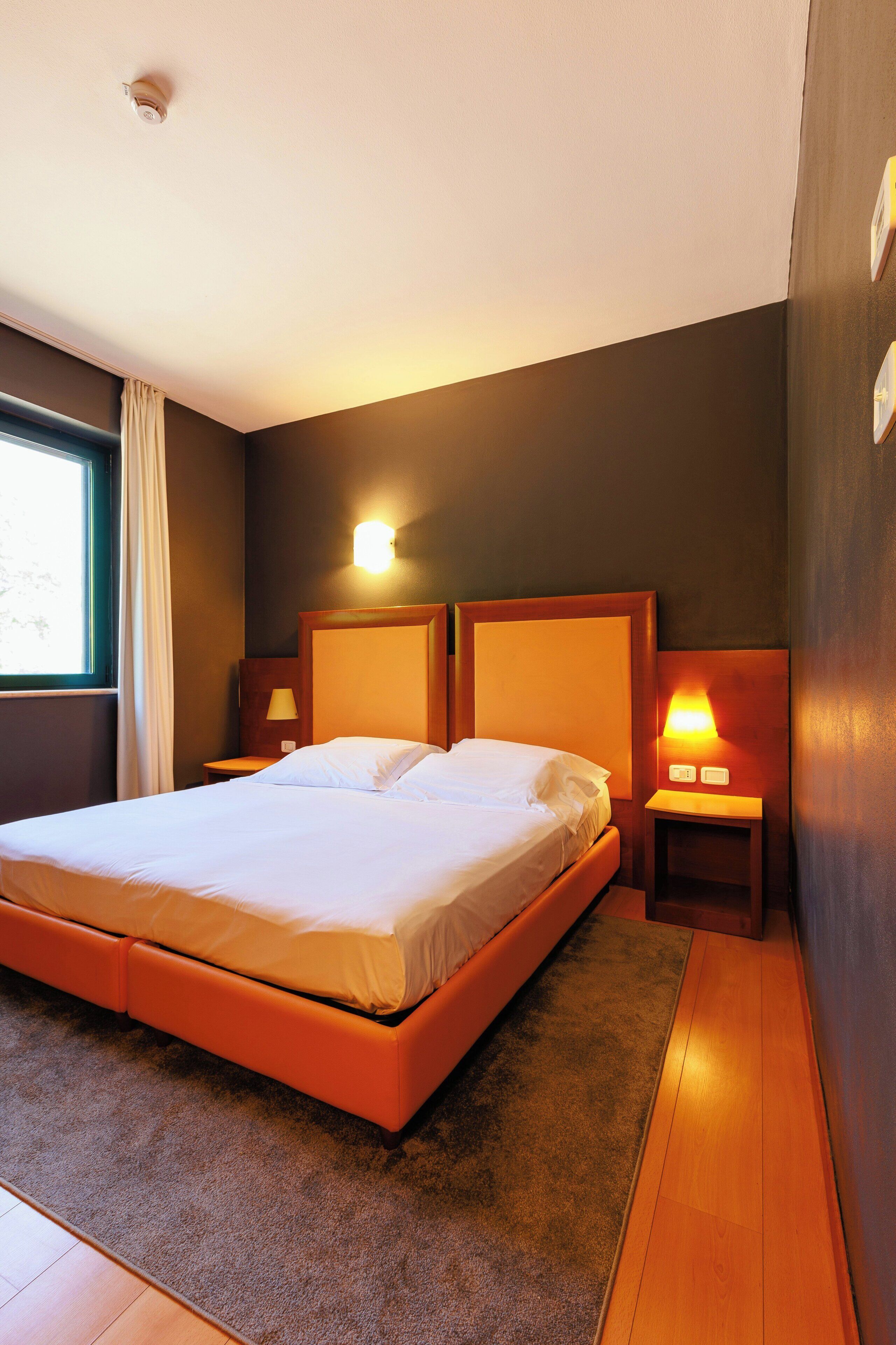 Quarto de hotel com uma cama grande, duas mesas de cabeceira, um criado-mudo, uma janela com cortinas e um candeeiro fixado na parede.