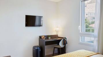 Quarto de hotel com cama, escrivaninha, cadeira, televisão, luminária e mala.