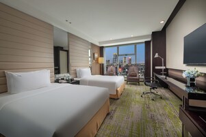 Quarto de hotel com duas camas, uma escrivaninha, uma cadeira, televisão e vista para a cidade.