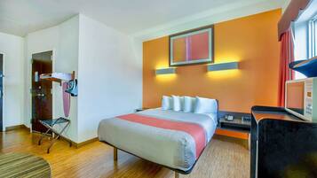 Quarto de hotel com cama, mesa de cabeceira e escrivaninha com cadeira. Há uma porta e uma janela com cortinas.