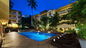Uma piscina com espreguiçadeiras e guarda-sóis, cercada por palmeiras e edifícios de hotel.