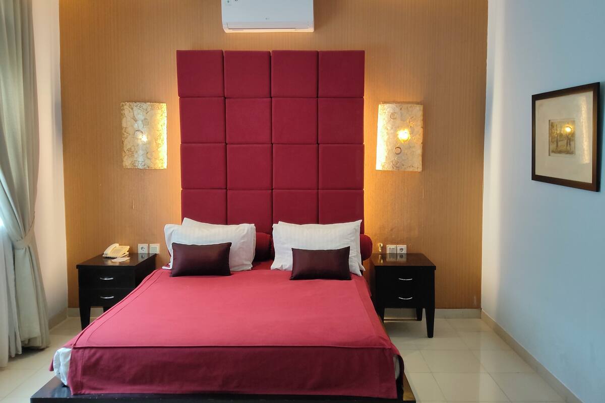 Quarto de hotel com uma cama grande, duas mesas de cabeceira, um aparelho de ar condicionado e um quadro na parede.