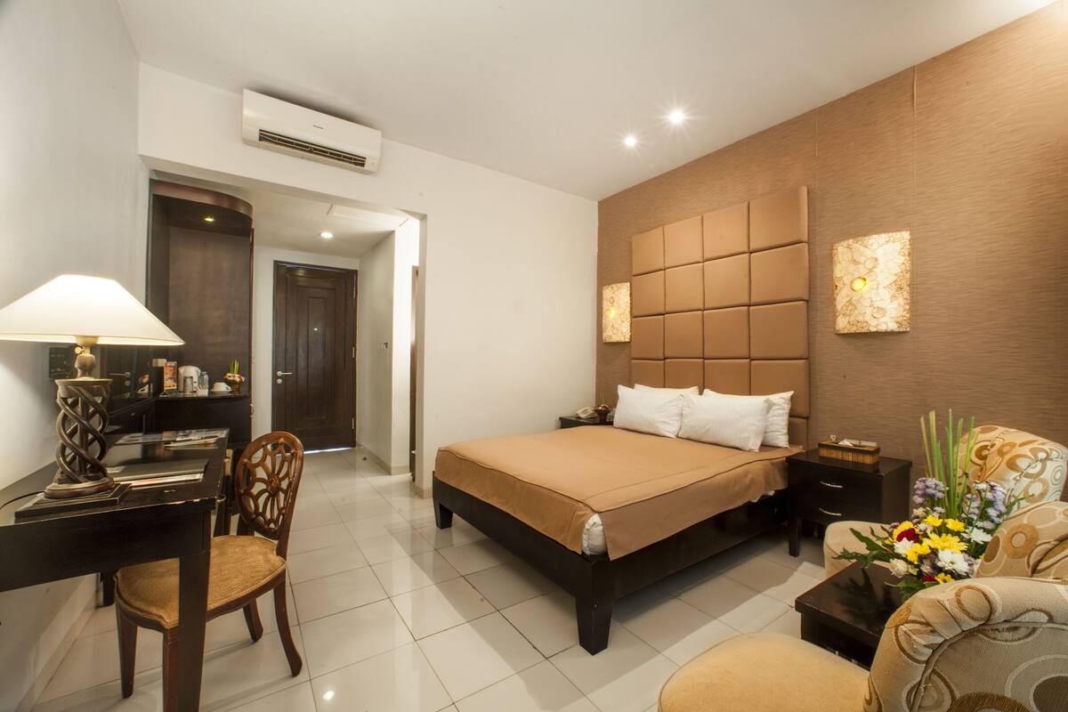 Quarto de hotel com cama, escrivaninha, cadeira, mesa de cabeceira e luminária na parede.