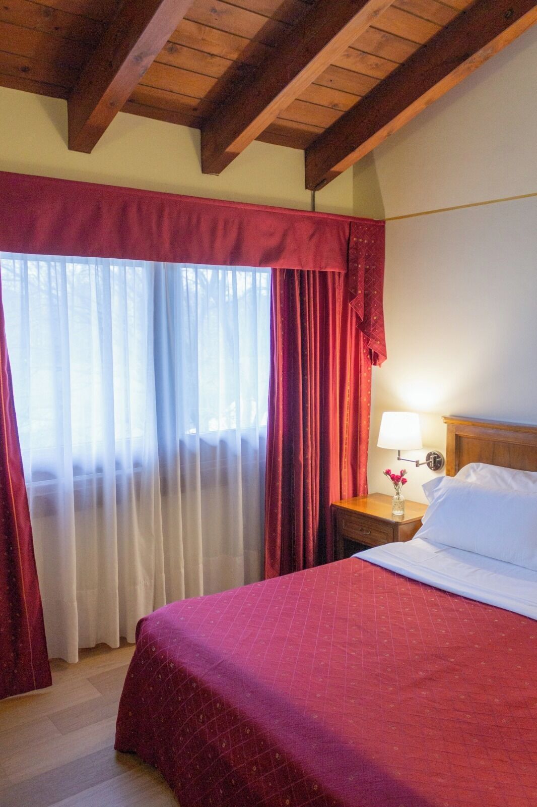 Quarto de hotel com cama, mesa de cabeceira com abajur e janela com cortinas.