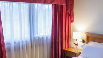 Quarto de hotel com cama, mesa de cabeceira com abajur e janela com cortinas.