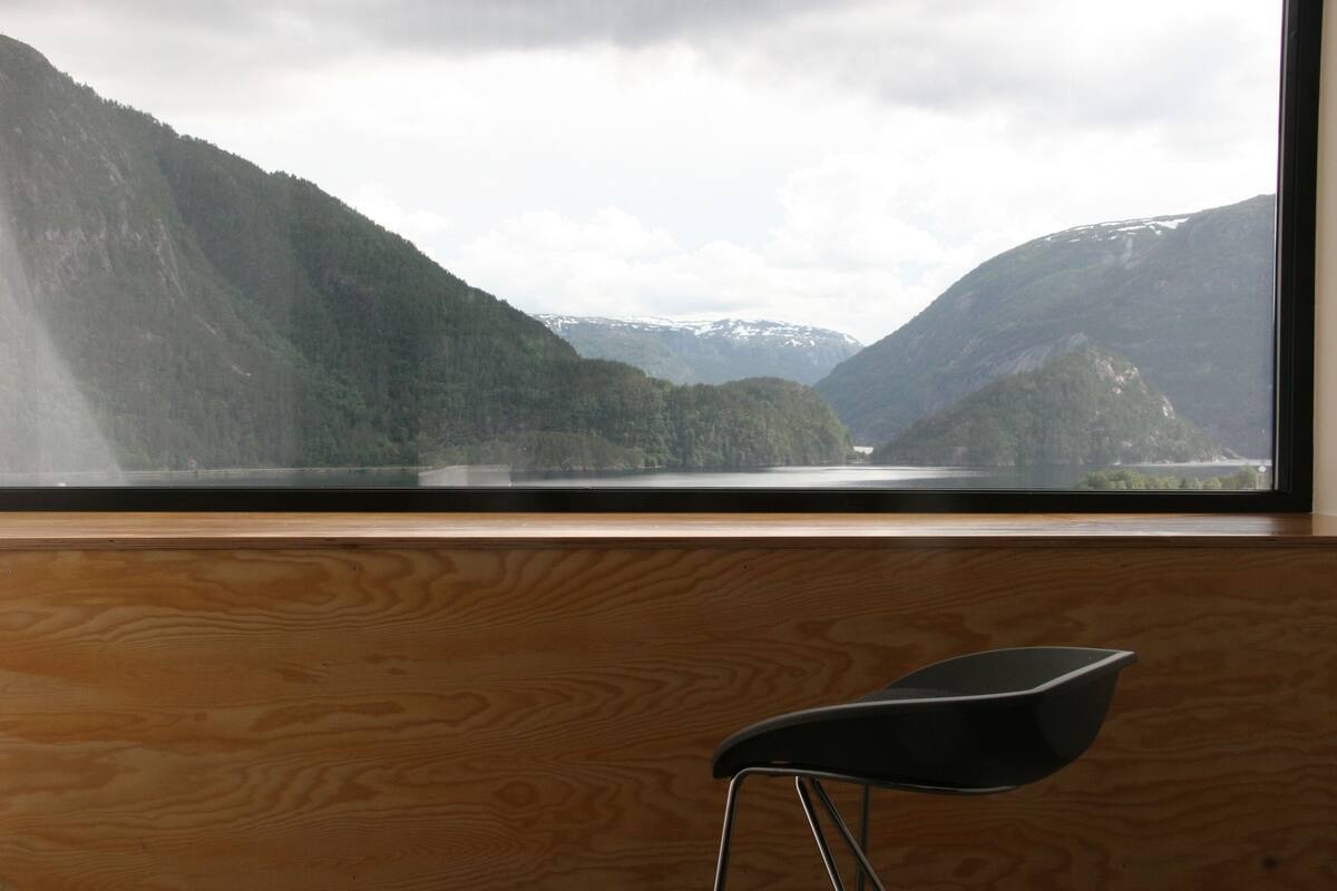 Vista da janela com interior de madeira, mostrando uma paisagem montanhosa com um lago.