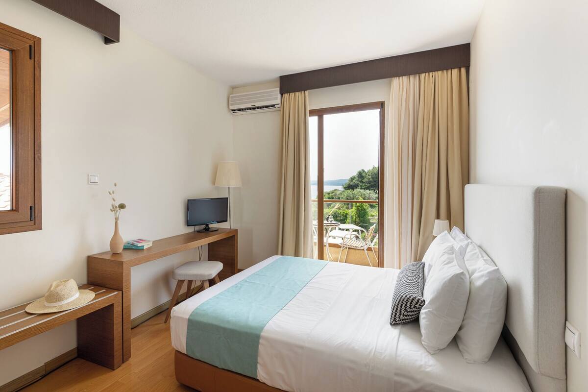Quarto de hotel com cama, escrivaninha, cadeira, TV e varanda com vista.