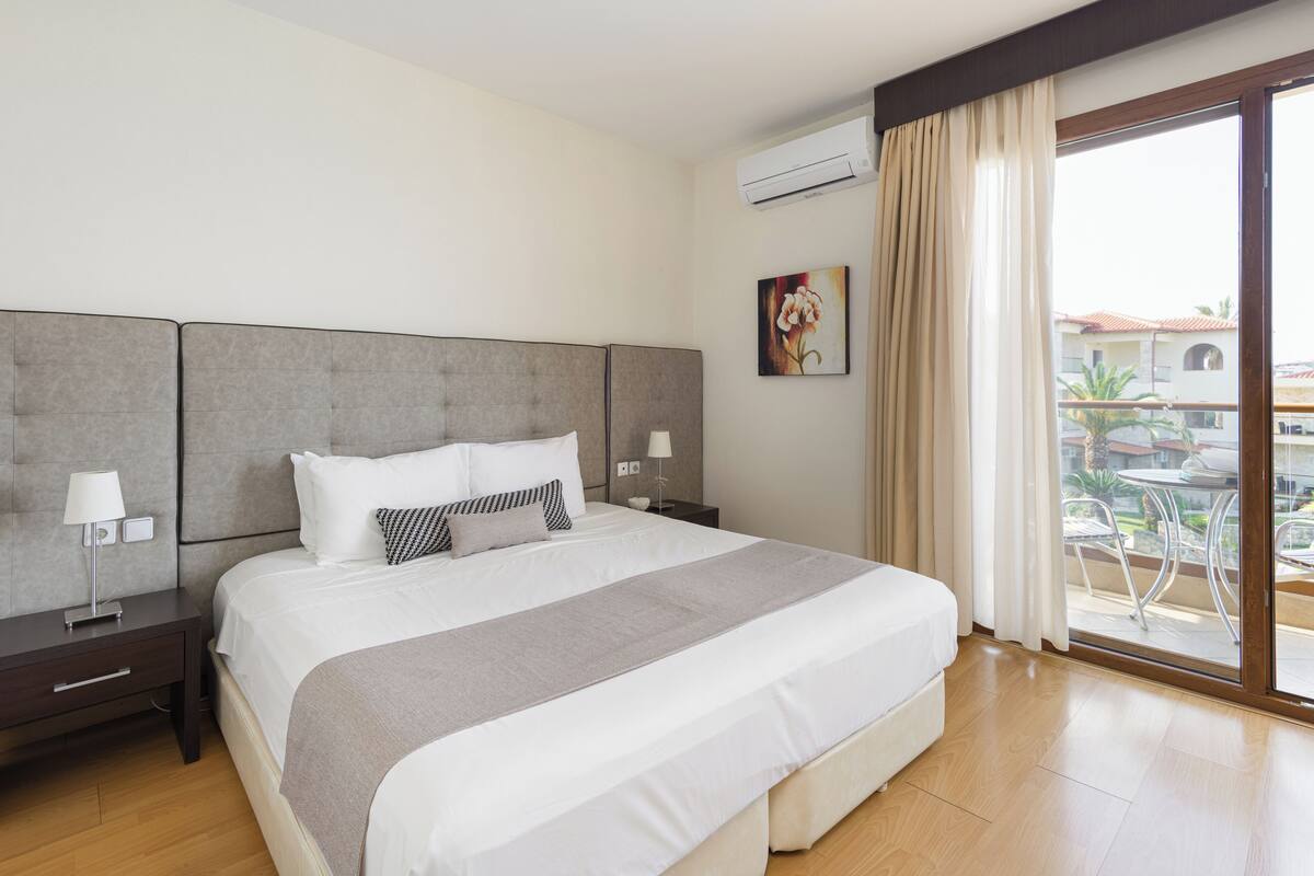 Quarto de hotel moderno com uma cama grande, mesa de cabeceira com abajur, janela com cortinas e vista para o exterior.
