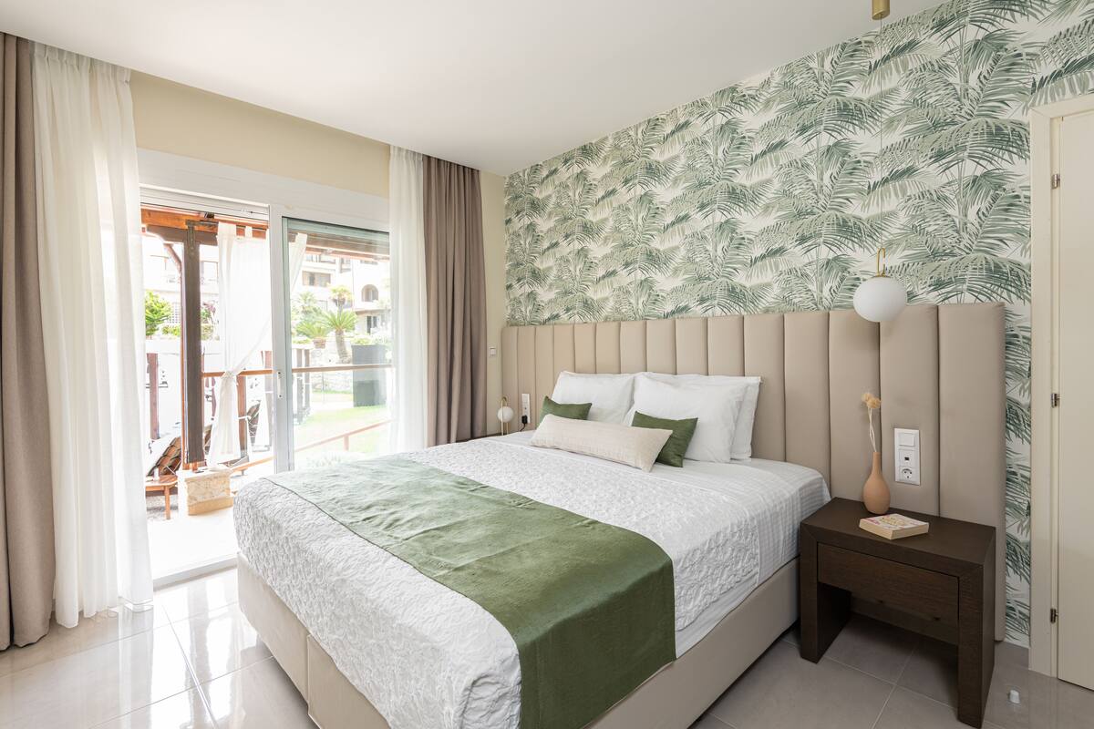 Quarto com cama grande, mesa de cabeceira, abajur e vista para o exterior.