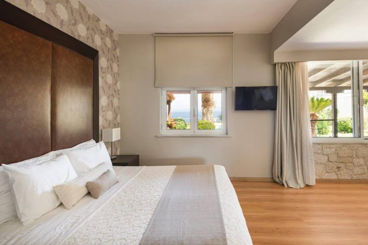 Quarto com cama grande, TV fixada na parede, janela com vista para área verde e parede de pedra.