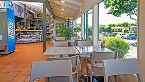Um restaurante com mesas internas, um balcão com diversos itens e vista para o estacionamento.