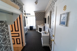 Quarto de hotel com papel de parede estampado, porta branca com número dourado, mesa branca pequena, espelho e quadro emoldurado na parede.