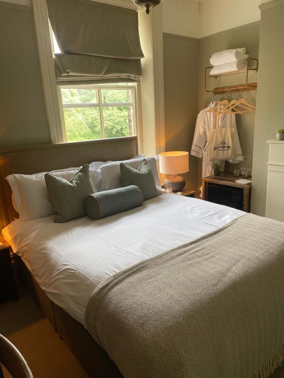 Uma cama bem arrumada com um manta estampada, uma mesa de cabeceira com luminária, uma janela com cortinas e um cabideiro com toalha e camisa.