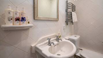 Casa de banho com lavatório branco, duas torneiras, um espelho, um porta-toalhas e uma sanita. Há três frascos de champô numa prateleira.