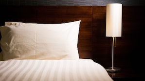 Uma cama bem arrumada com lençóis brancos e um único travesseiro branco, uma mesa de cabeceira com abajur e uma cabeceira de madeira escura.