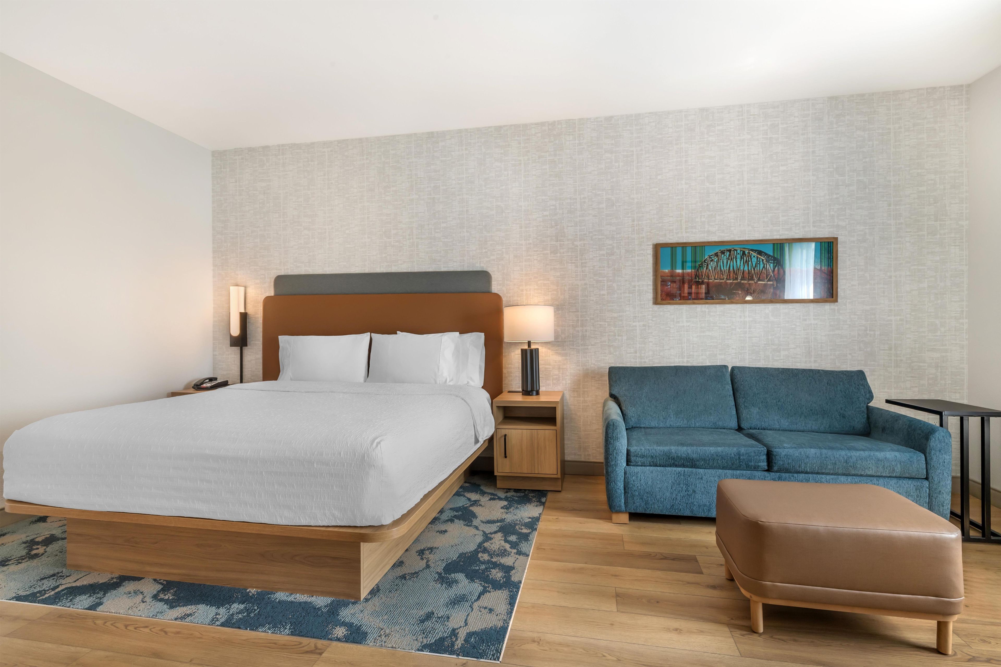 Quarto de hotel com cama, sofá azul, um banquinho, mesa de cabeceira, abajur e um quadro na parede.