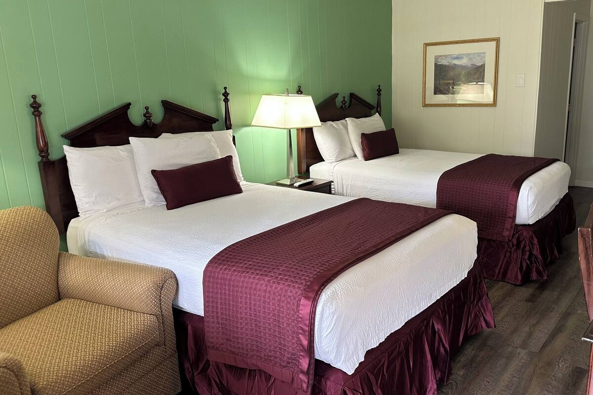 Quarto de hotel com duas camas, cada uma com lençóis brancos e colchas bordô, uma cabeceira de madeira, mesa de cabeceira com abajur e um quadro na parede.