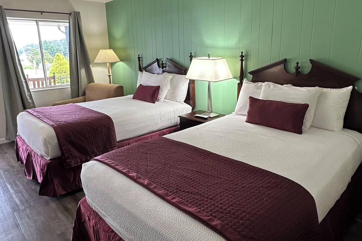 Quarto de hotel com duas camas, paredes verdes, cabeceiras de madeira e uma janela grande com cortinas.