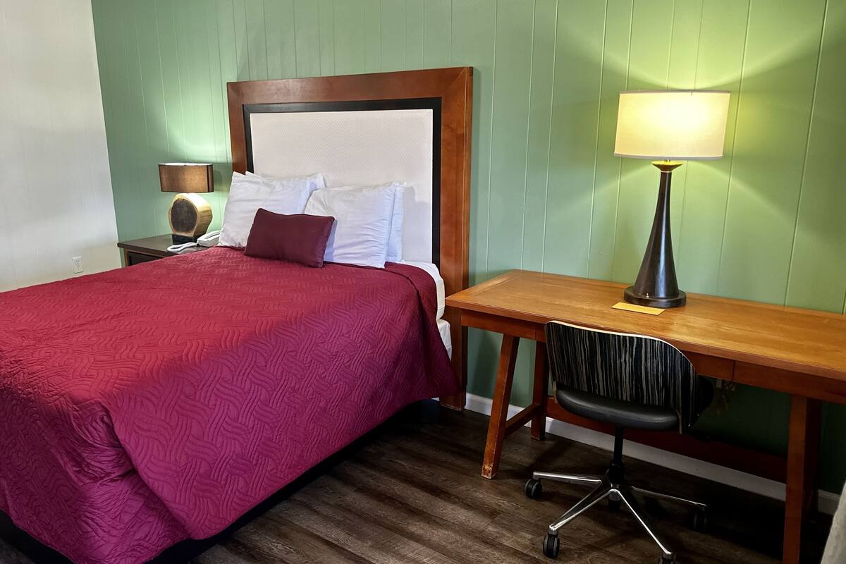 Quarto de hotel com cama, escrivaninha, cadeira, abajur e telefone.