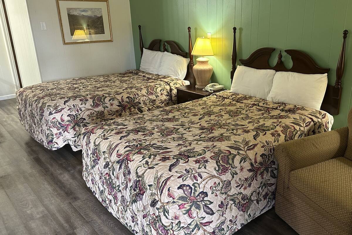 Quarto de hotel com duas camas, uma mesa de cabeceira com abajur, uma cadeira e um quadro na parede.