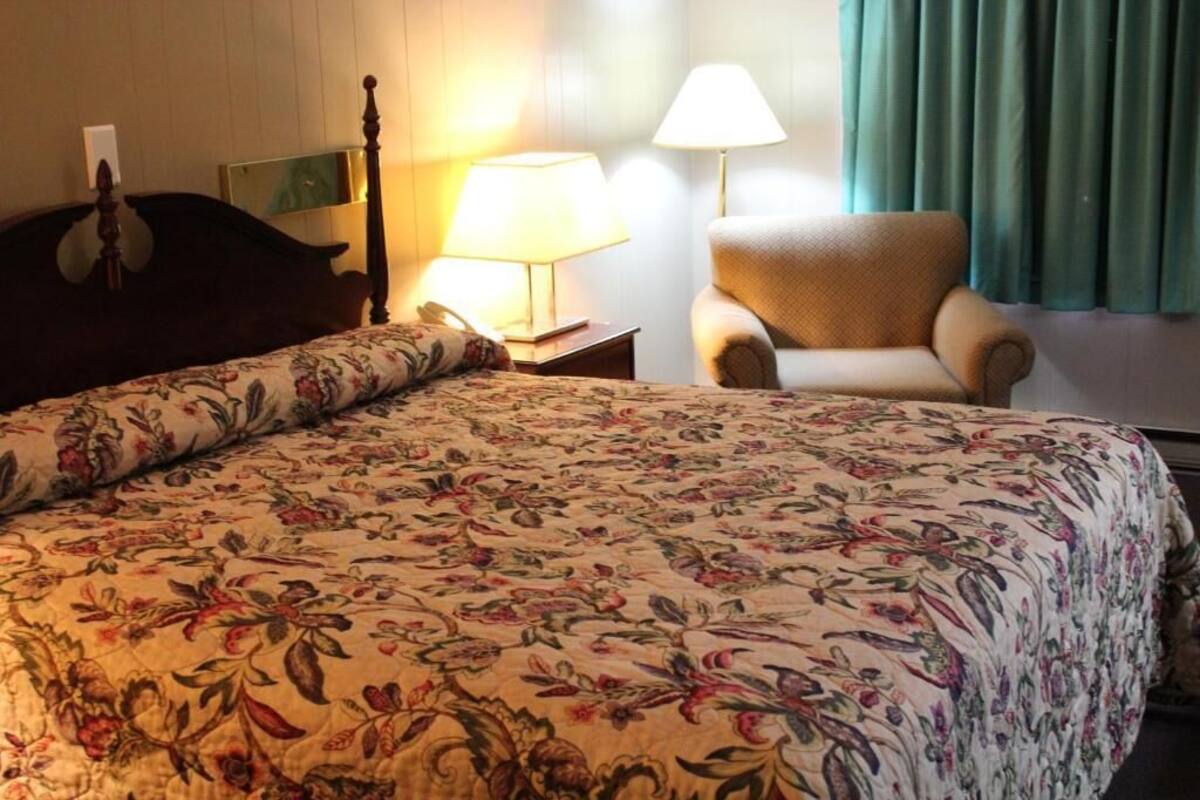 Cama bem arrumada com colcha florida, cabeceira de madeira, luminária de cabeceira, cadeira e mesa de cabeceira com abajur.
