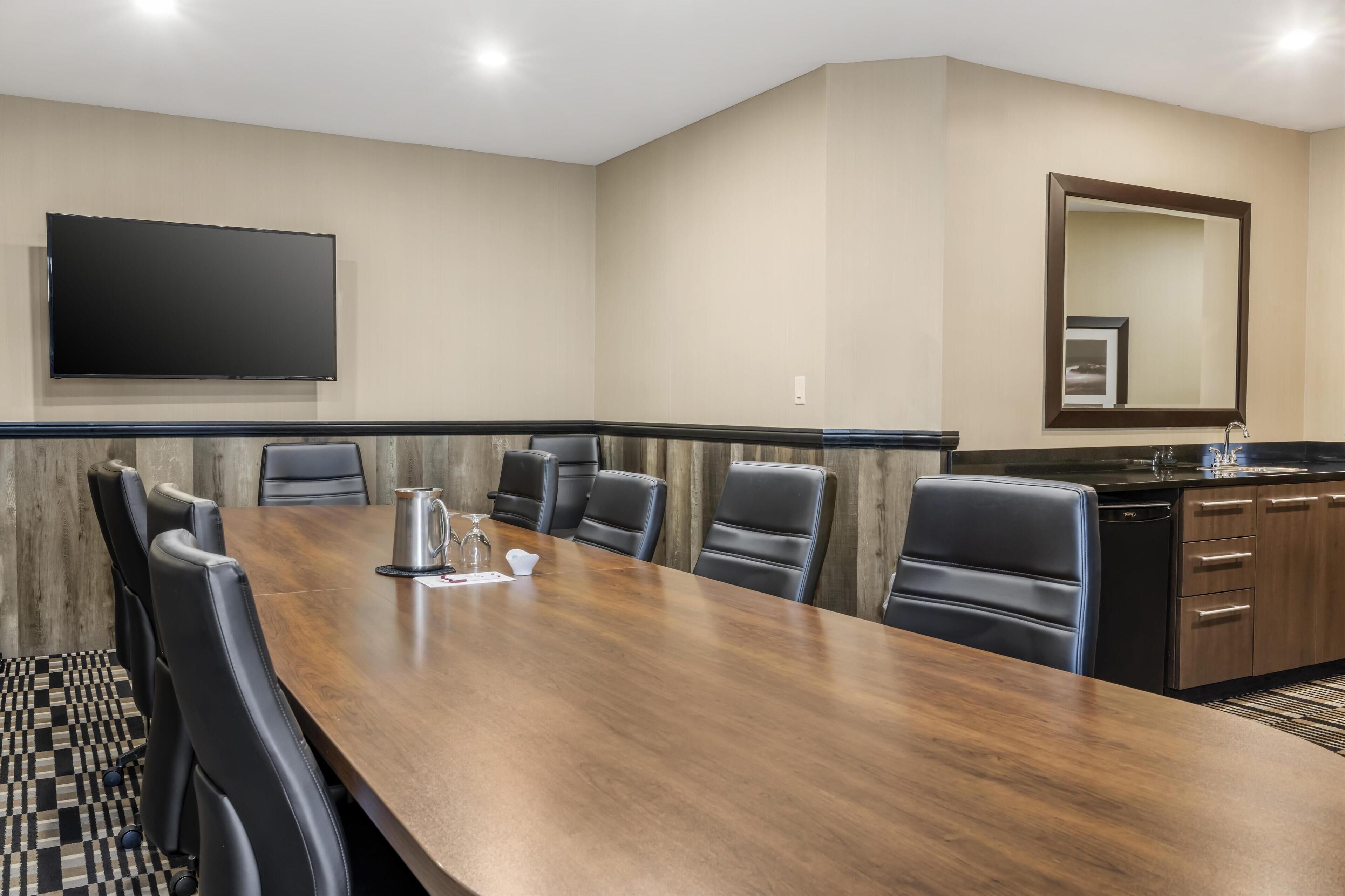 Uma sala de conferência com uma mesa comprida, cadeiras de couro preto, uma TV de tela plana, uma área de cozinha com pia e armários, e um tapete estampado.
