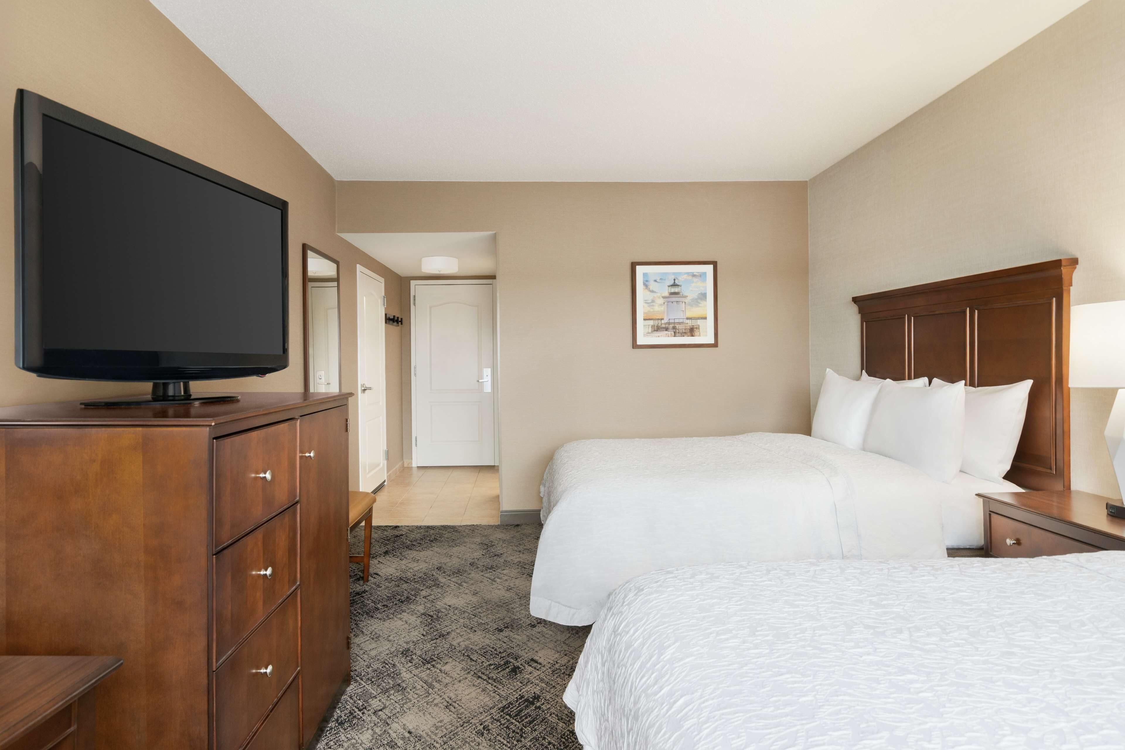 Quarto de hotel com televisão de tela plana sobre uma cômoda de madeira, duas camas com lençóis brancos, um quadro na parede e uma cabeceira de madeira.
