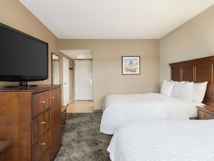 Quarto de hotel com televisão de tela plana sobre uma cômoda de madeira, duas camas com lençóis brancos, um quadro na parede e uma cabeceira de madeira.