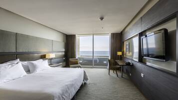 Quarto de hotel com uma cama grande, uma escrivaninha com cadeira, televisão de tela plana na parede e uma janela ampla com vista para o oceano.