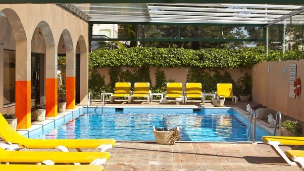 Área da piscina com cadeiras de descanso amarelas, piscina transparente e área coberta com arcos.
