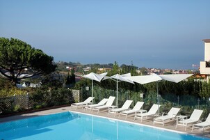 Piscina com espreguiçadeiras brancas e guarda-sóis, cercada por vegetação e com vista para uma cidade costeira.