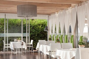 Uma área de refeições ao ar livre com mesas e cadeiras, um lustre pendurado e vista para o verde através de grandes janelas.