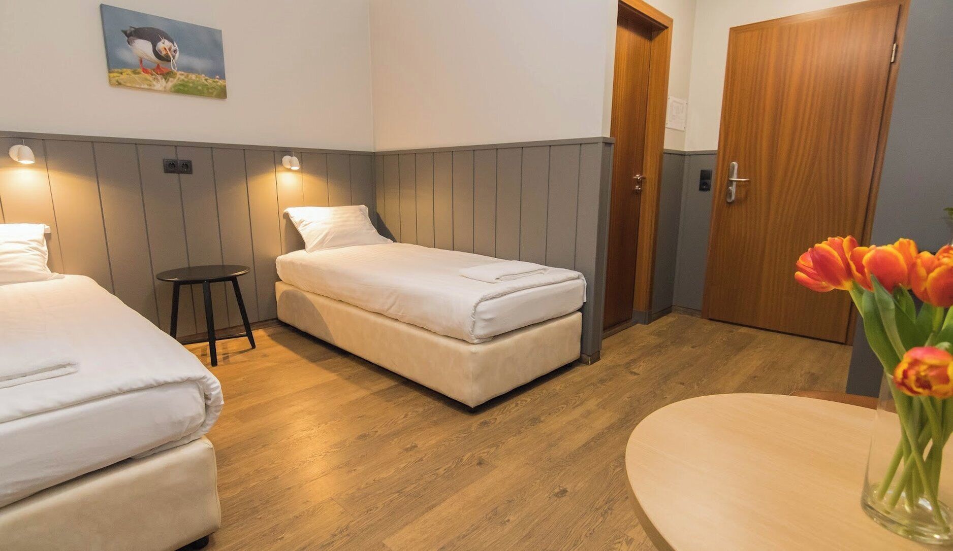Quarto de hotel com duas camas, uma mesinha redonda, uma porta de madeira e um ponto de luz fixado na parede.