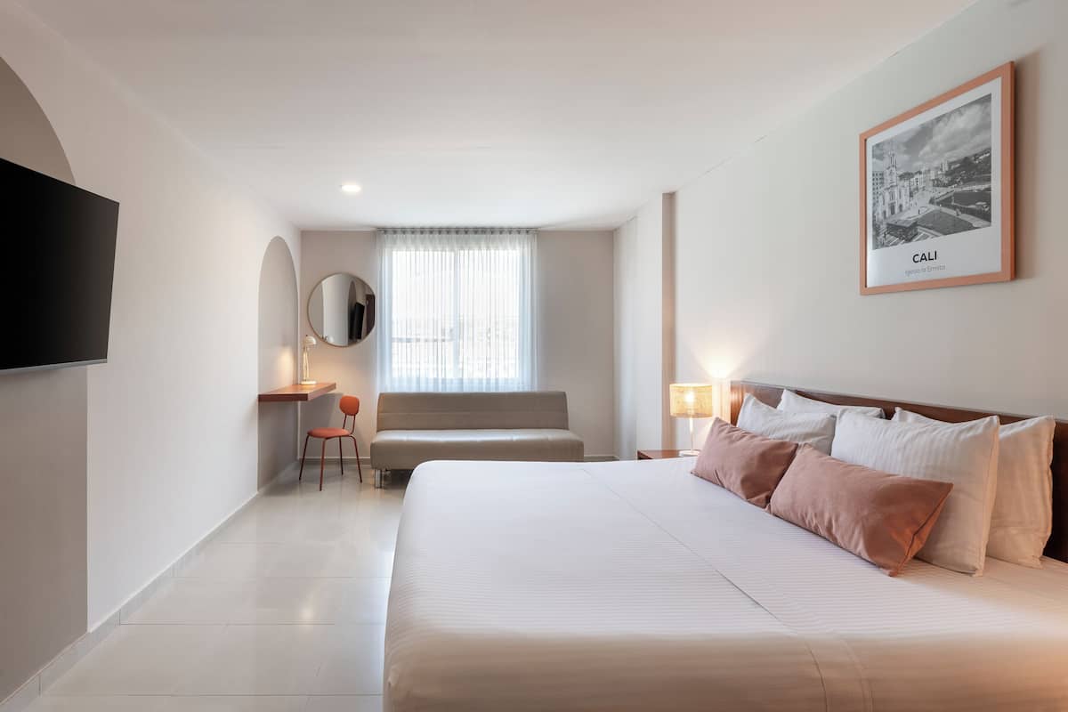 Quarto de hotel com uma cama grande, uma escrivaninha com cadeira, TV fixada na parede e um quadro com uma vista urbana.