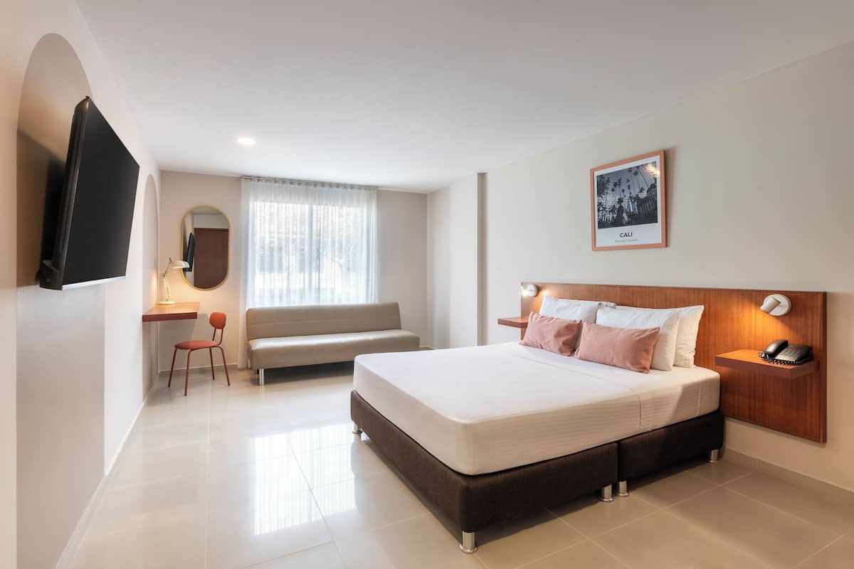 Quarto de hotel moderno com uma cama grande, uma escrivaninha com cadeira, uma televisão e um quadro de uma palmeira na parede.