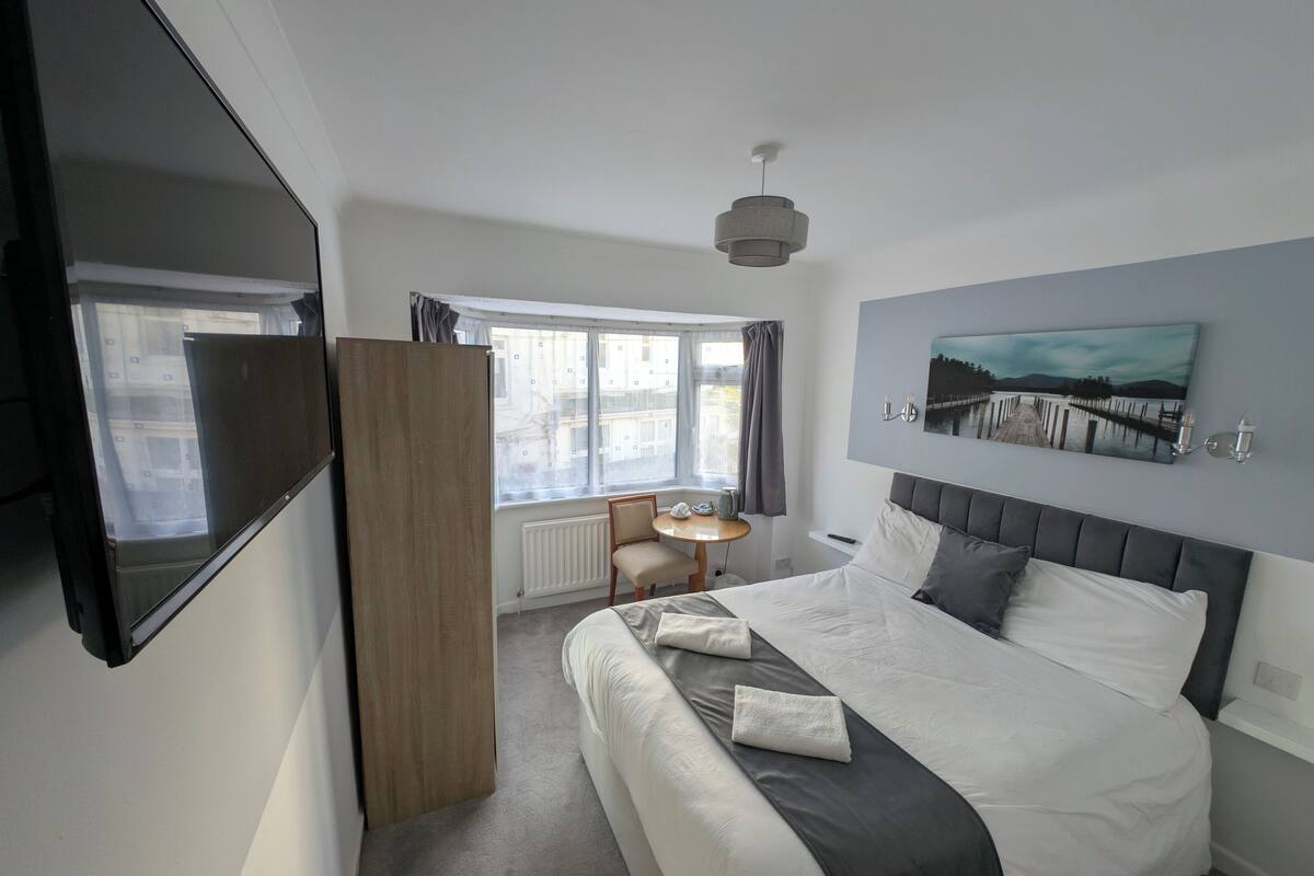 Quarto de hotel moderno com uma cama grande, uma escrivaninha com cadeira, uma janela com cortinas e um quadro na parede.