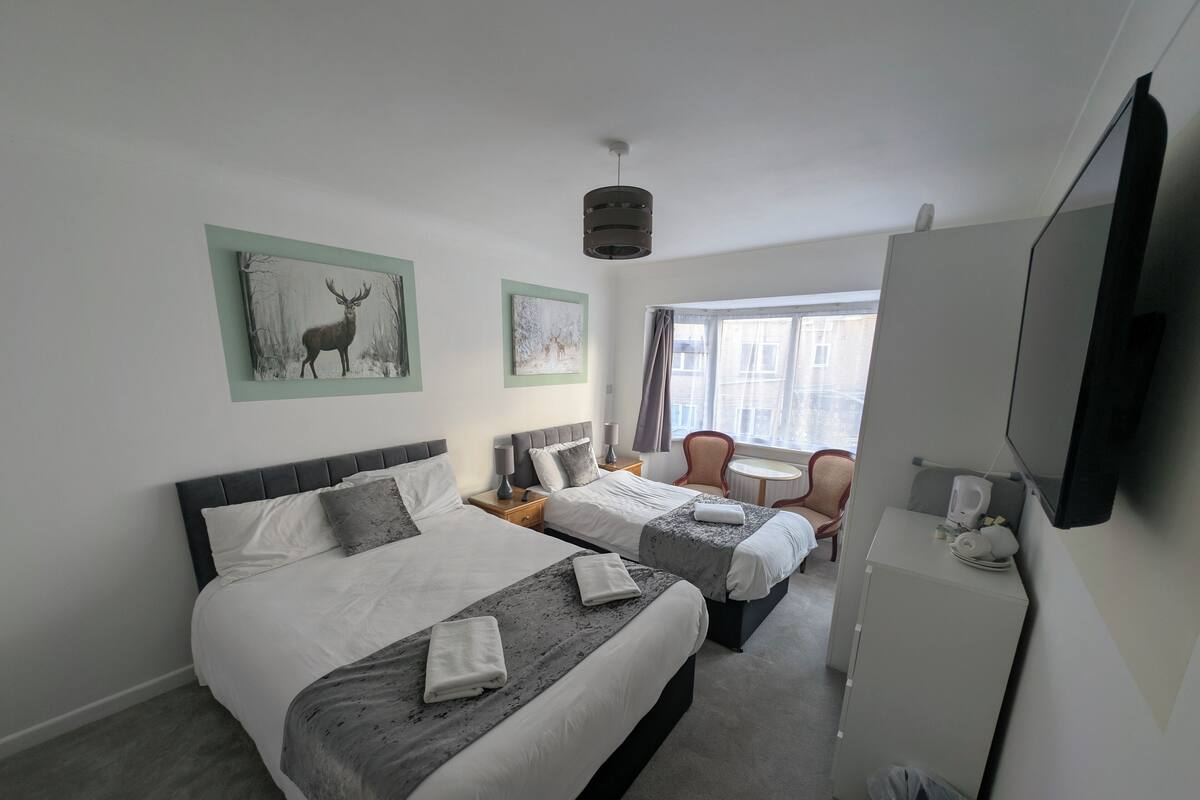 Quarto de hotel com duas camas, uma mesinha com cadeiras, televisão e obras de arte nas paredes.