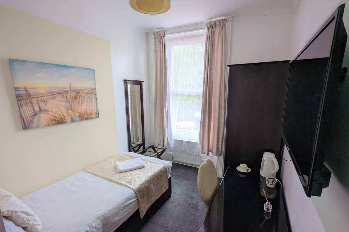 Quarto de hotel com cama, escrivaninha, cadeira, espelho, quadro e janela com cortinas.