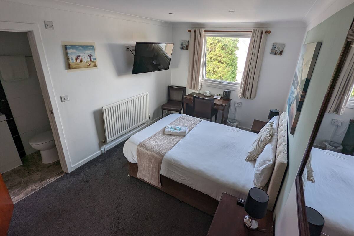 Quarto de hotel com cama, escrivaninha, cadeira, televisão, janela com cortinas e radiador.