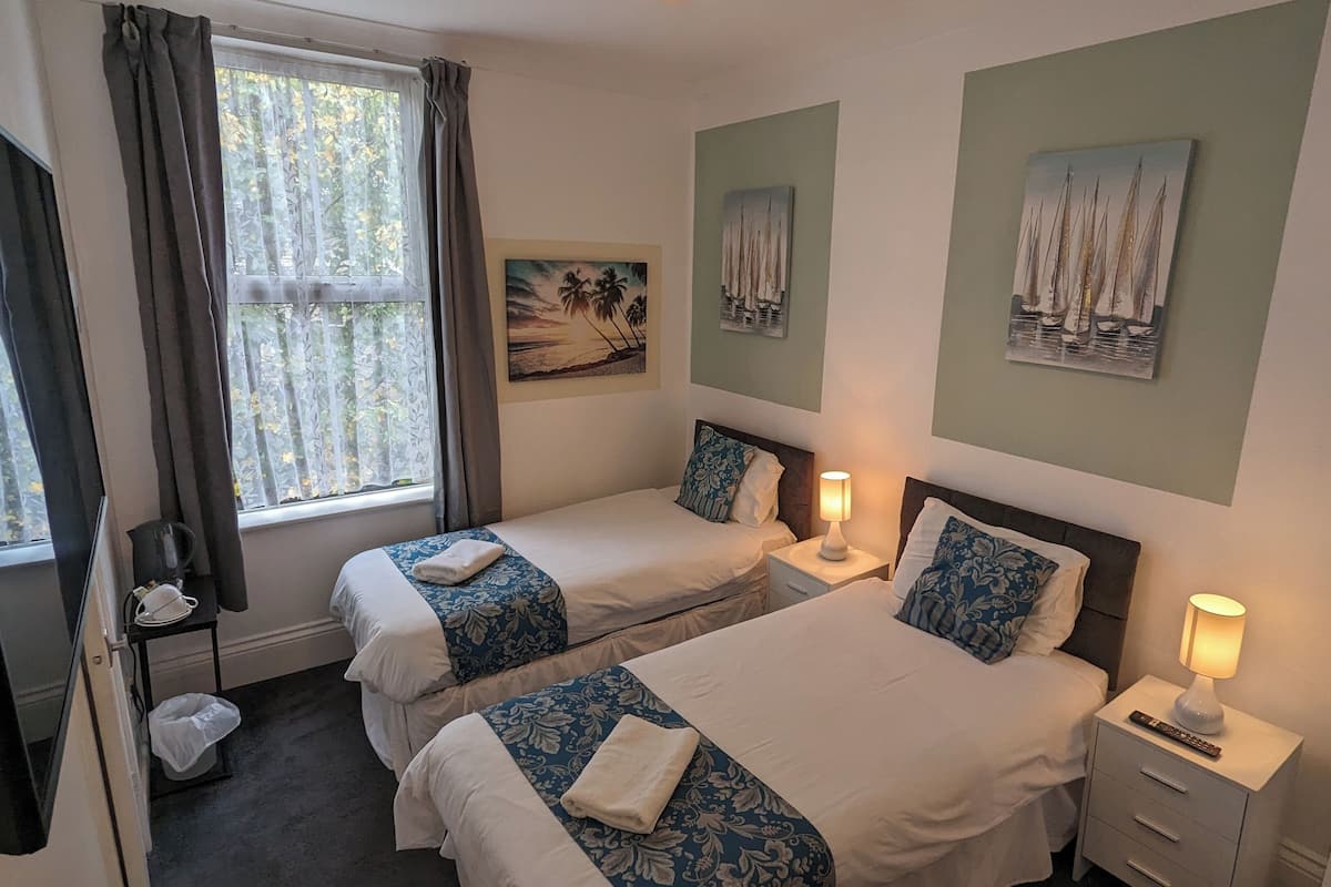 Quarto de hotel com duas camas, janela com cortinas, pintura de veleiros e luminária na mesa de cabeceira.